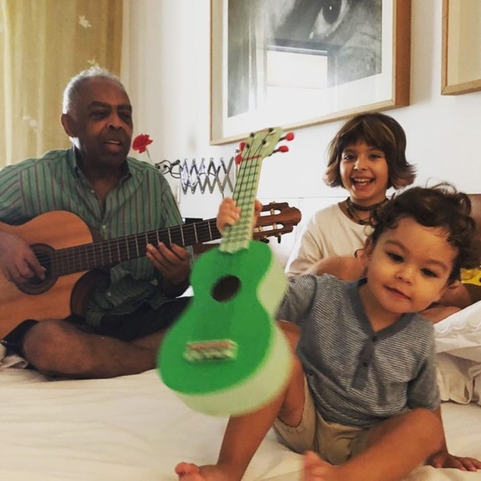 Gilberto Gil posa na cama tocando violão com filhos de Bela Gil